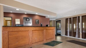 A hotel reception area with a wooden reception desk, a sign reading "Quality by Choice Hotels," and a green doormat.
