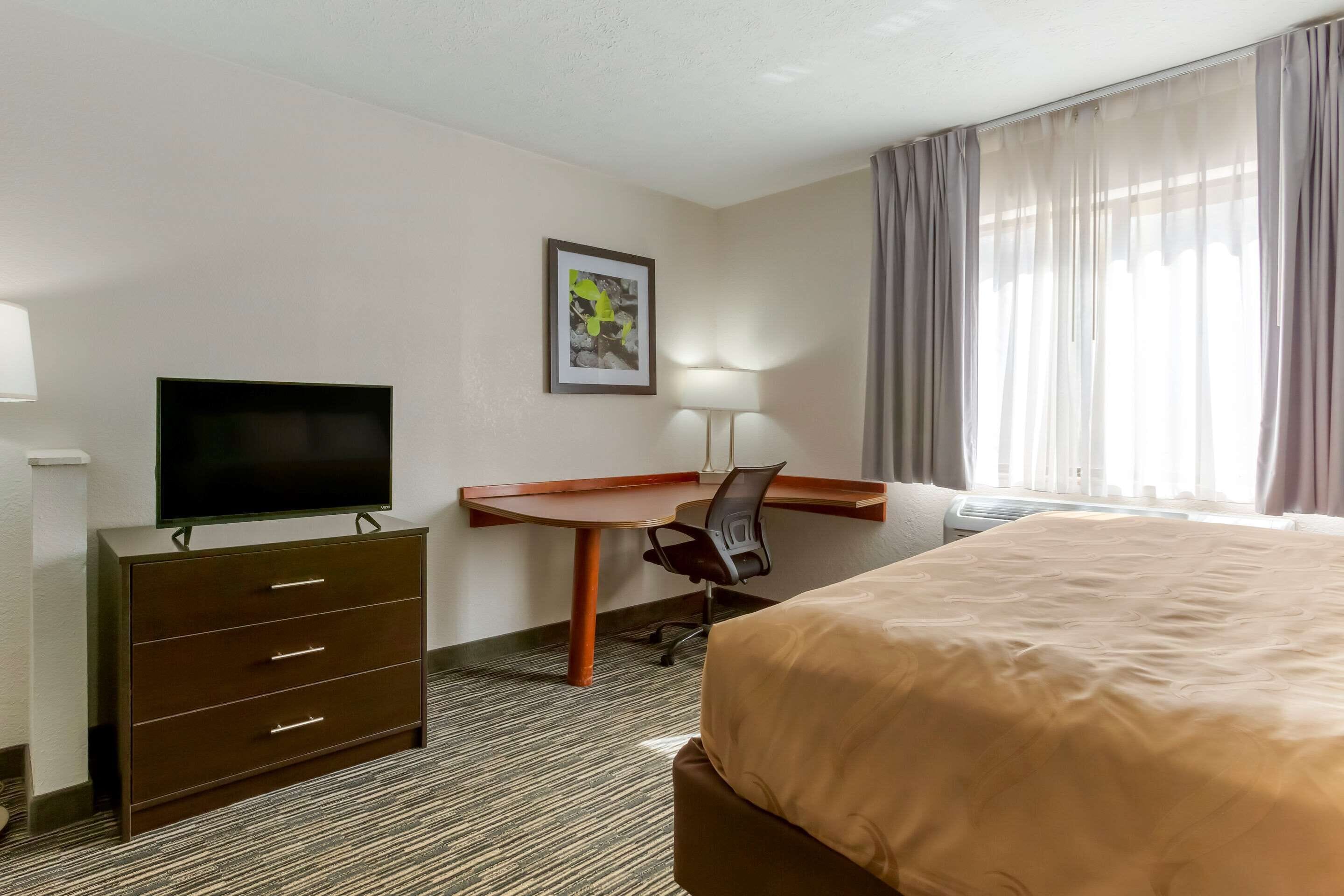 Quarto de hotel com cama, escrivaninha, cadeira, televisão e abajur.