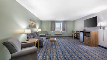 Quarto de hotel com sofá, poltrona, mesa de centro, televisão de tela plana, mini-geladeira, micro-ondas e uma grande janela com cortinas.