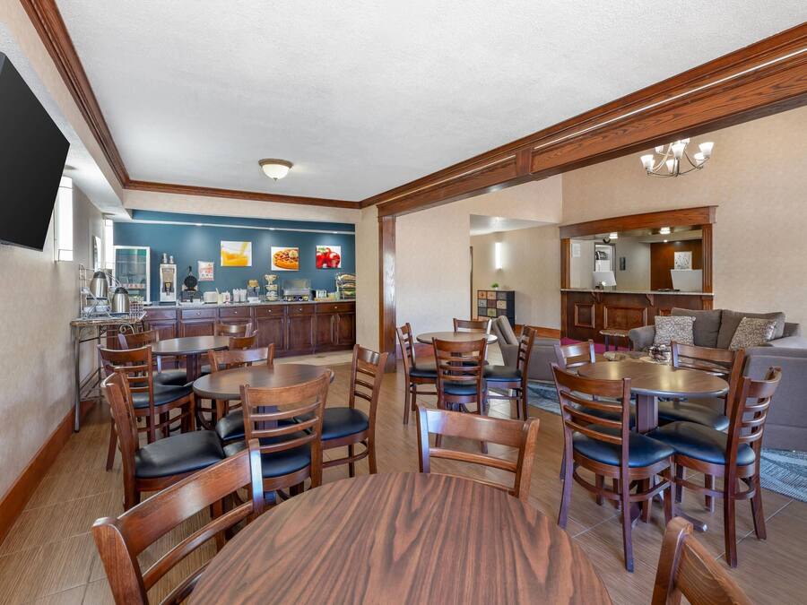 Uma sala de jantar com mesas e cadeiras de madeira, uma cozinha ao fundo, e uma sala de estar com sofá e televisão na parede.