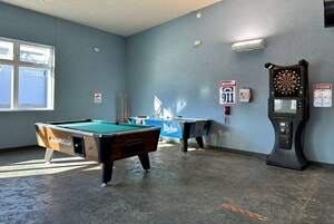 Mesa de bilhar com superfície de feltro azul, alvo de dardos e mesa de sinuca em um espaço com paredes azuis e uma janela.