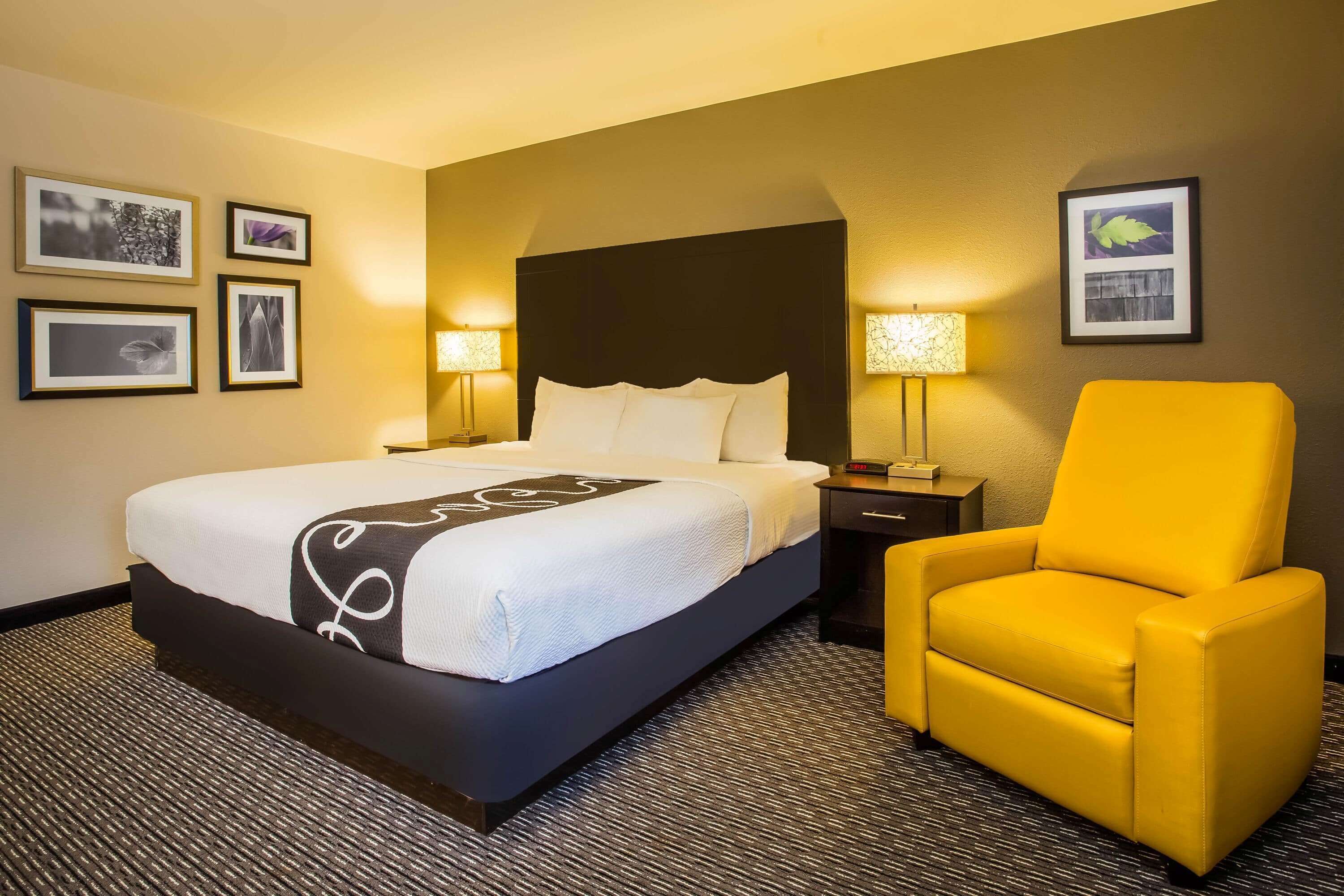 Quarto de hotel com uma cama grande, duas mesas de cabeceira, uma poltrona amarela e quadros nas paredes.