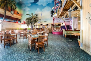 Restaurante com tema tropical, apresentando mesas e cadeiras de madeira, um palco e um aviso de 'Zona de Silêncio'.