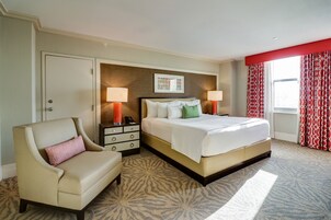 Quarto de hotel com uma cama, uma cadeira, uma mesinha de cabeceira e uma janela com cortinas.