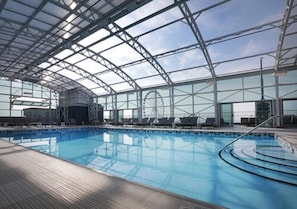 Piscina interna com teto de vidro, cadeiras de descanso e uma grande janela.