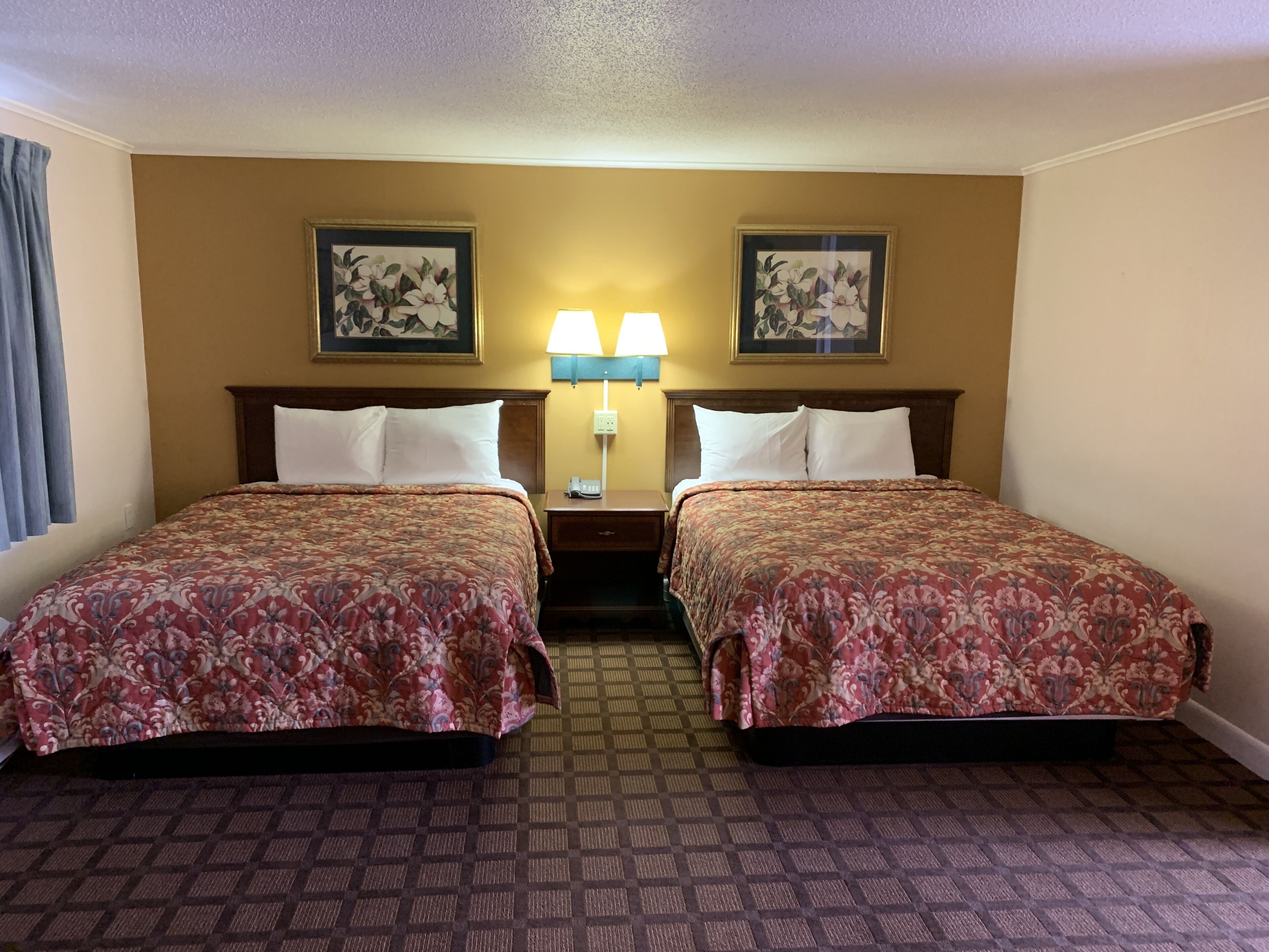 Quarto de hotel com duas camas, uma mesa de cabeceira com luminária e arte floral na parede.