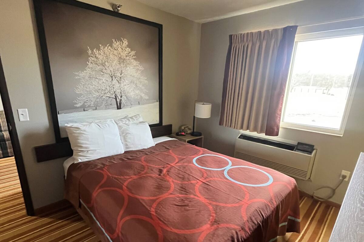 Quarto de hotel com cama, mesa de cabeceira, abajur, janela com cortinas e uma obra de arte pendurada na parede.