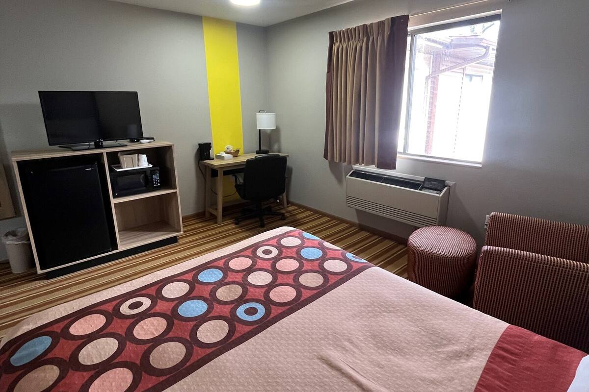 Quarto de hotel com cama, escrivaninha, televisão, micro-ondas e janela com cortinas.