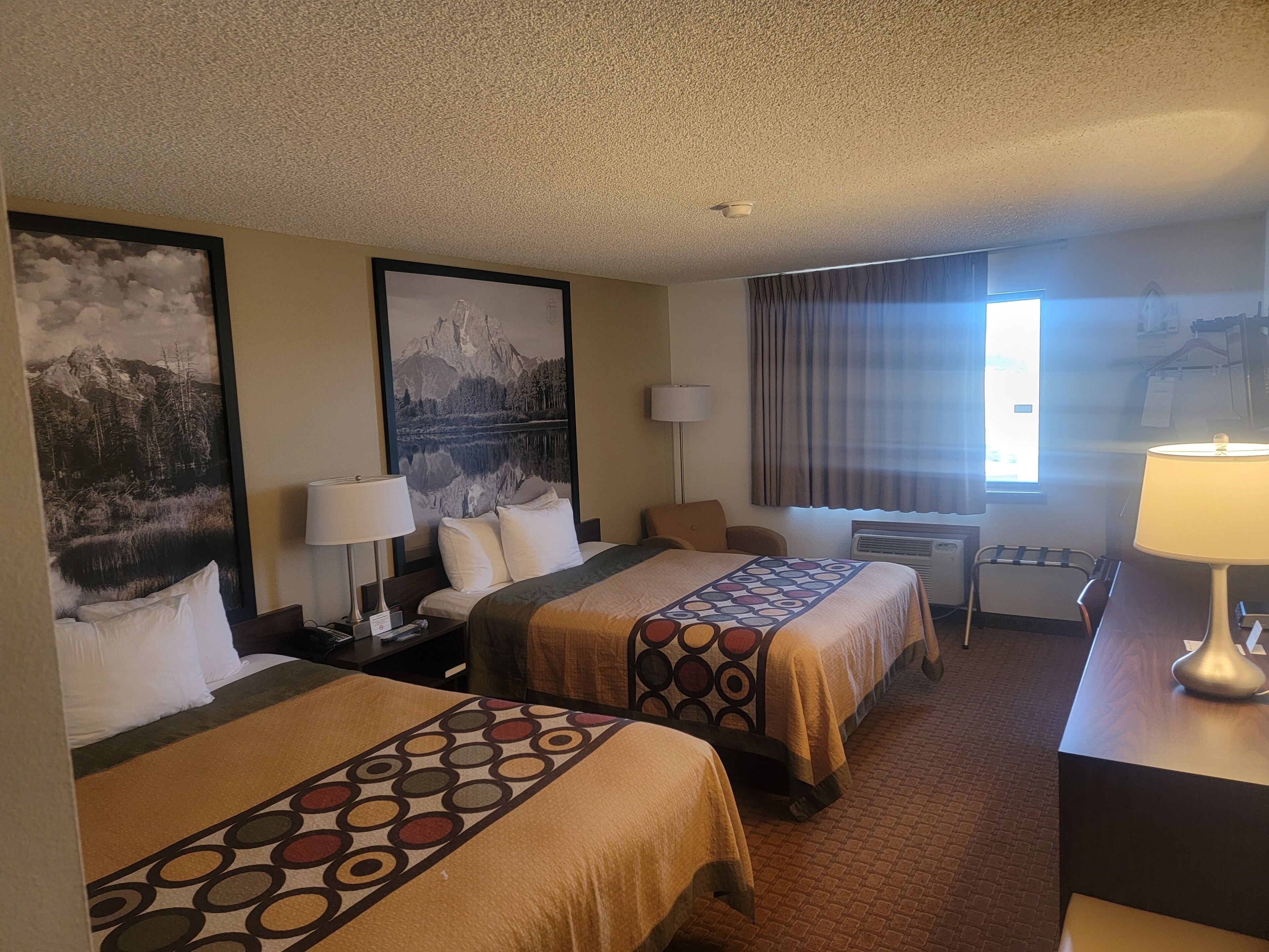 Quarto de hotel com duas camas, uma escrivaninha, uma cadeira, um abajur e uma janela com cortinas.