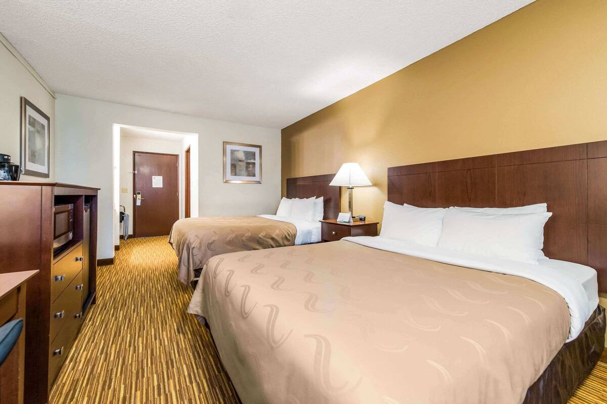 Quarto de hotel com duas camas, uma cabeceira de madeira, uma escrivaninha com luminária e piso coberto por carpete.