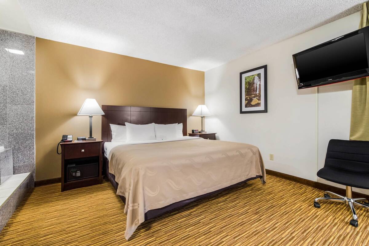 Quarto de hotel com cama, duas luminárias laterais, uma televisão, uma cadeira e uma mesinha de centro com telefone e cofre.