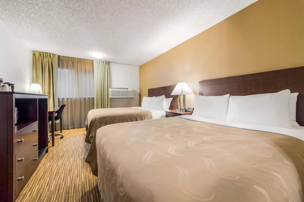 Quarto de hotel com duas camas, uma escrivaninha, uma cadeira, um abajur e um ar condicionado.