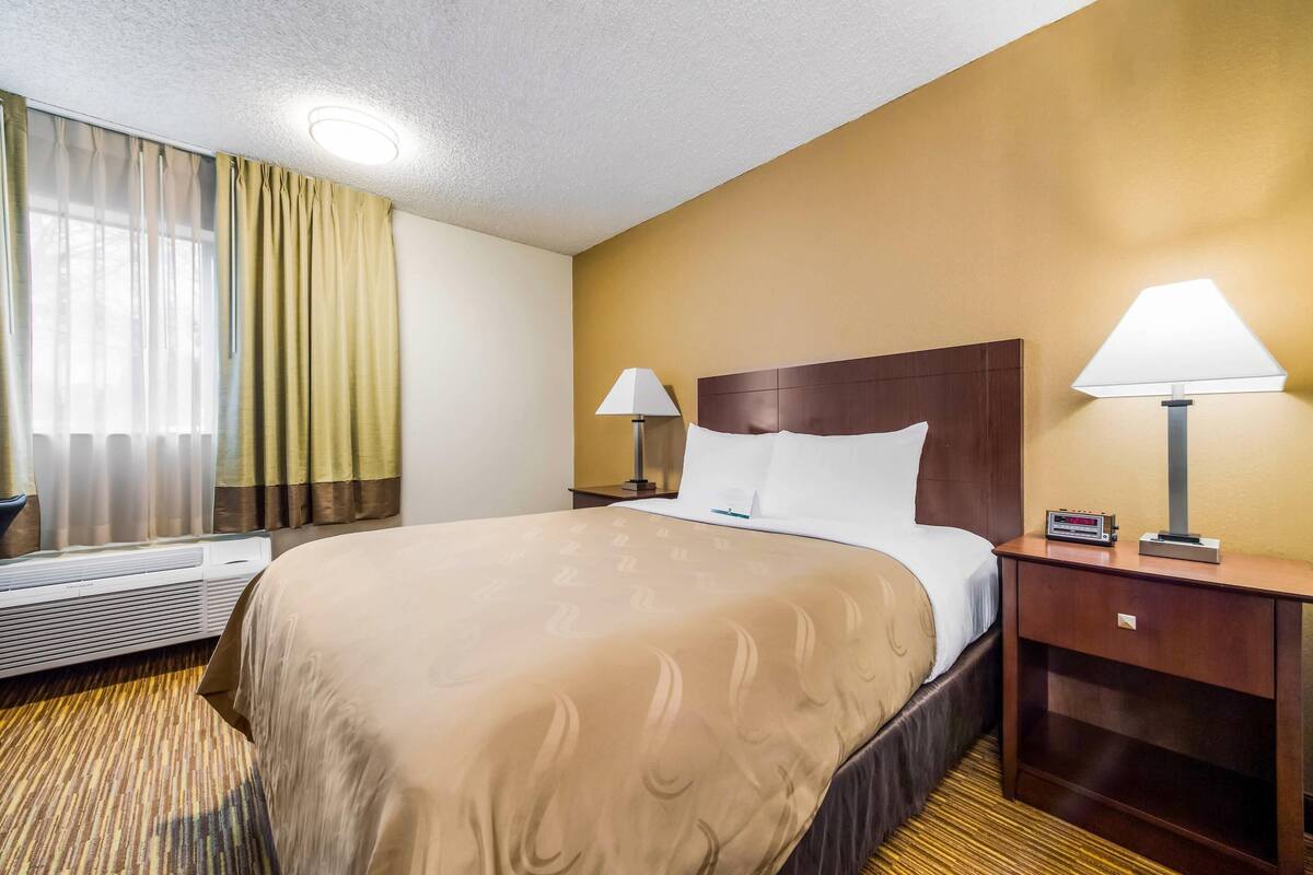 Quarto de hotel com uma cama grande, duas mesas de cabeceira com abajures, um aquecedor e uma janela com cortinas.