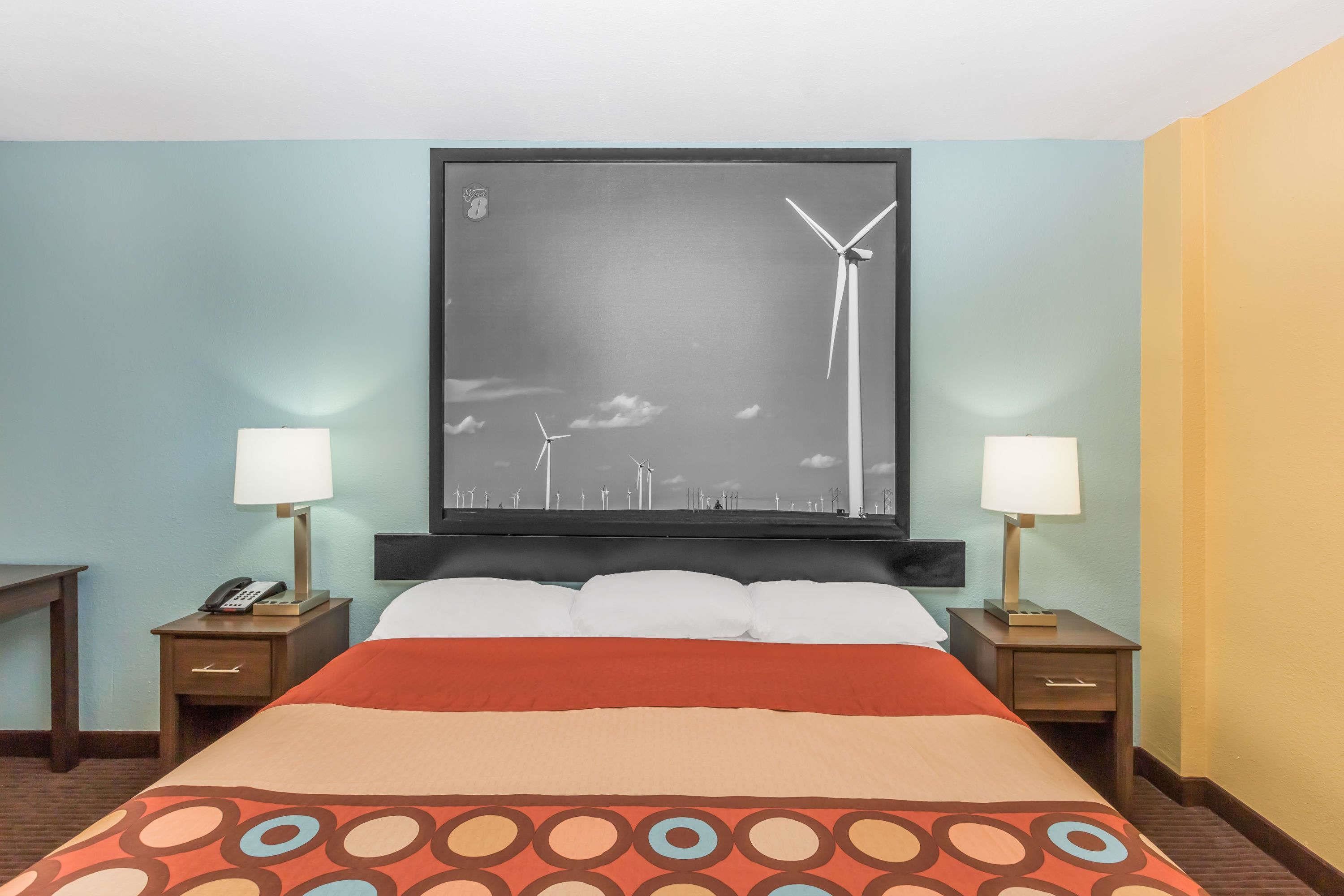 Quarto de hotel com cama, duas mesas de cabeceira com luminárias, uma obra de arte na parede retratando turbinas eólicas e uma vista da cidade.