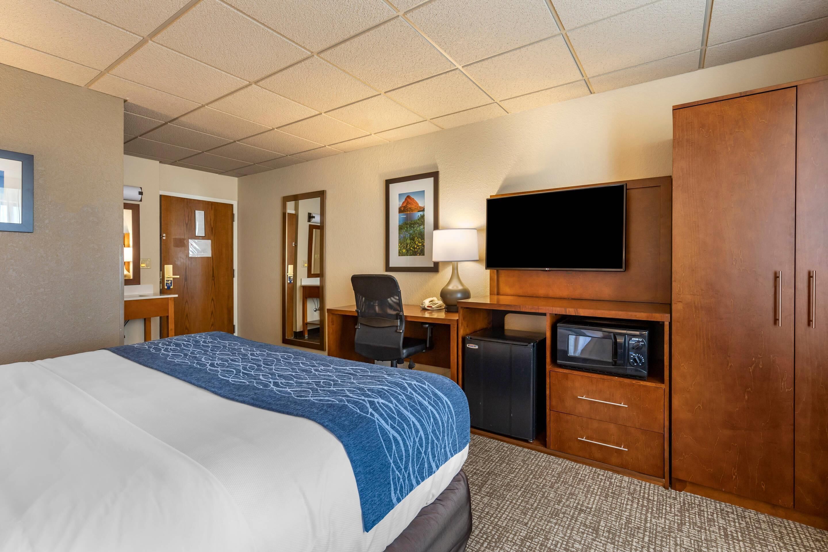 Quarto de hotel com uma cama grande, uma escrivaninha com cadeira, televisão, micro-ondas e armários de madeira.