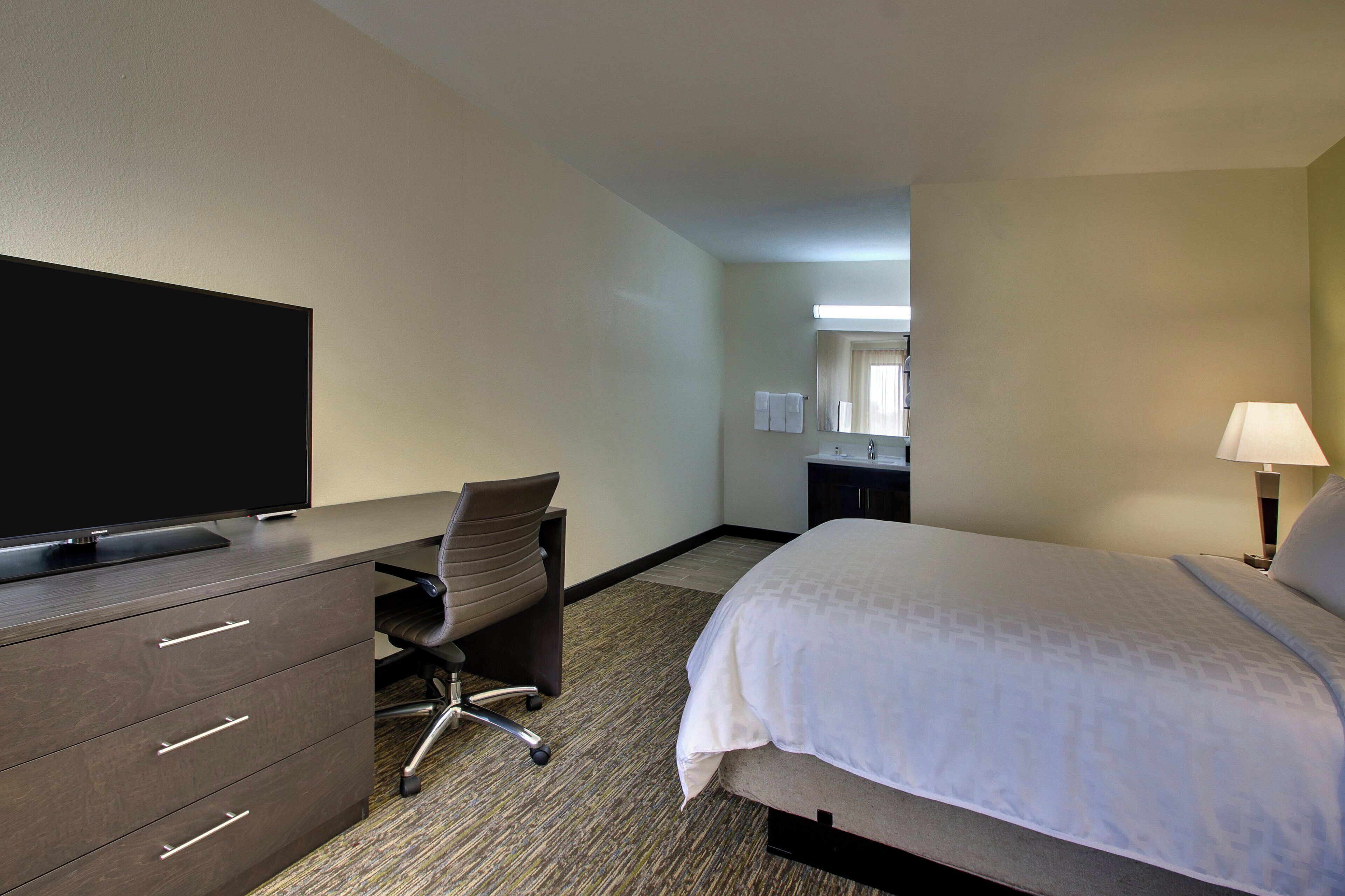 Quarto de hotel com cama, escrivaninha, cadeira, televisão, abajur e espelho.
