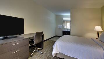 Quarto de hotel com cama, escrivaninha, cadeira, televisão, abajur e espelho.