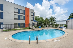 Uma piscina com escada de metal, cercada por grade e adjacente a um edifício de vários andares com fachada azul e laranja.