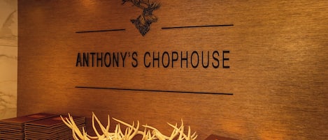 Pared de madeira com o texto "ANTHONY'S CHOPHOUSE" e um emblema de cabeça de veado. Há pilhas de cardápios e um arranjo decorativo de galhos de veado sobre a mesa.