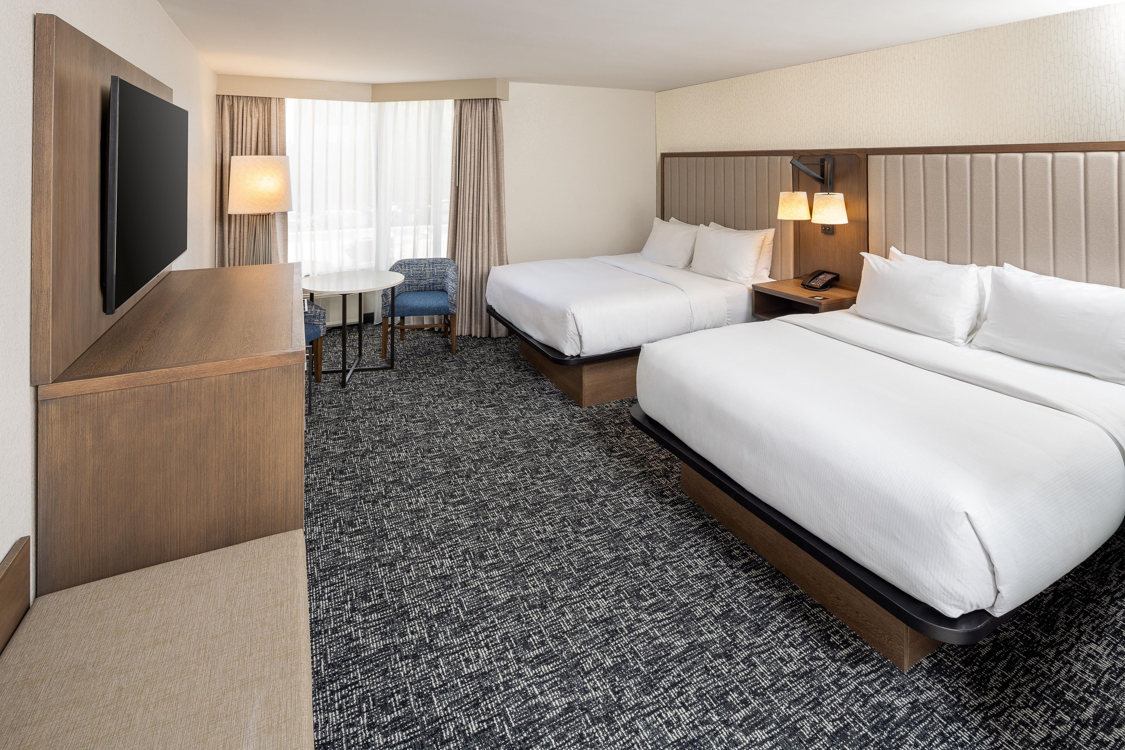 Quarto de hotel com duas camas, uma televisão de tela plana, uma mesa de trabalho com cadeira e uma janela com cortinas.