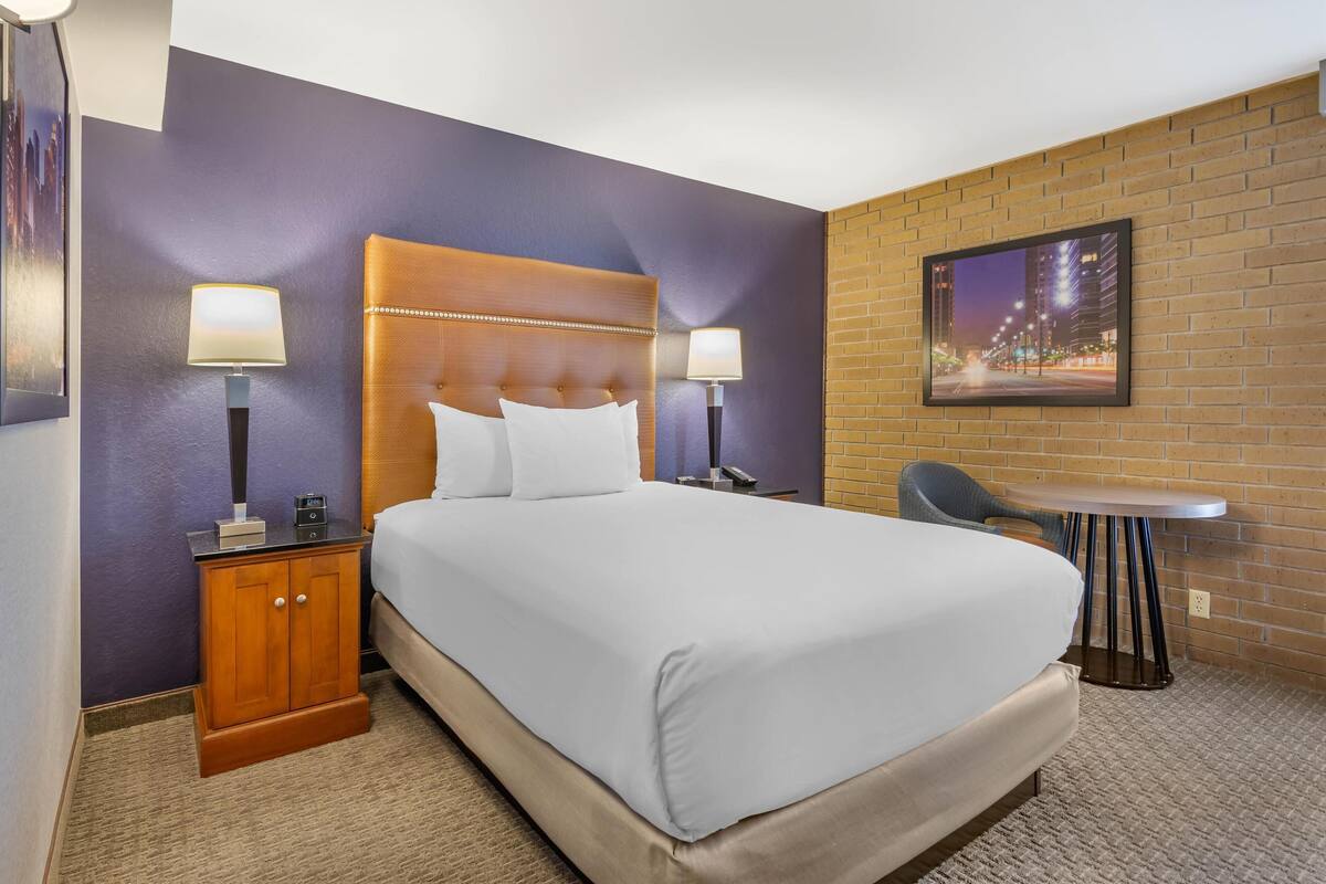 Quarto de hotel com uma cama grande, duas mesas de cabeceira com luminárias, uma mesa redonda pequena e uma pintura de paisagem urbana na parede.