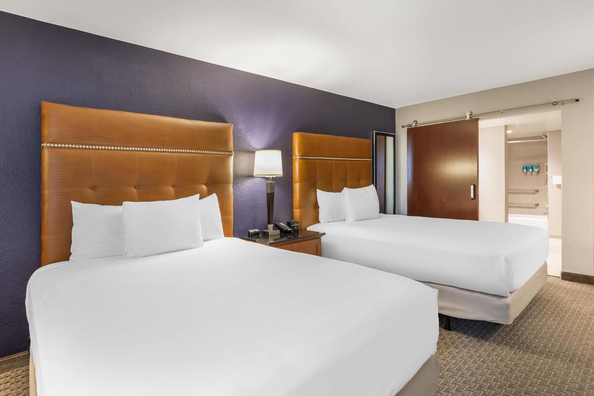 Quarto de hotel com duas camas, mesa de cabeceira, luminária e porta de correr para o banheiro.