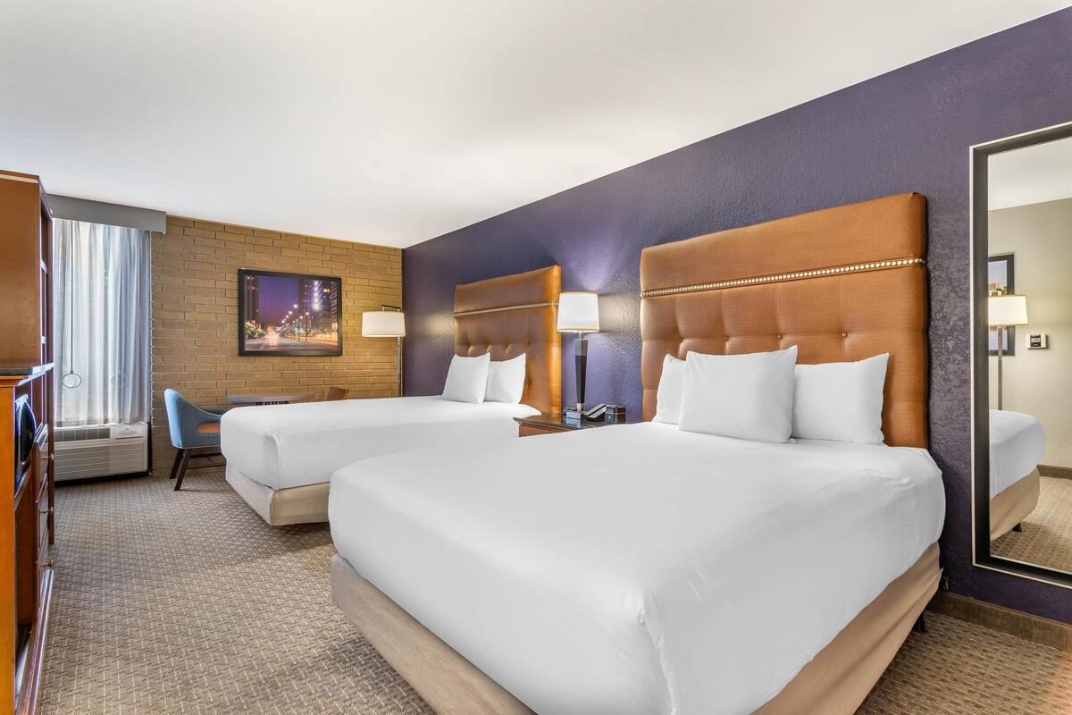 Quarto de hotel com duas camas, uma mesa, uma cadeira, um espelho grande e uma imagem de uma paisagem urbana na parede.