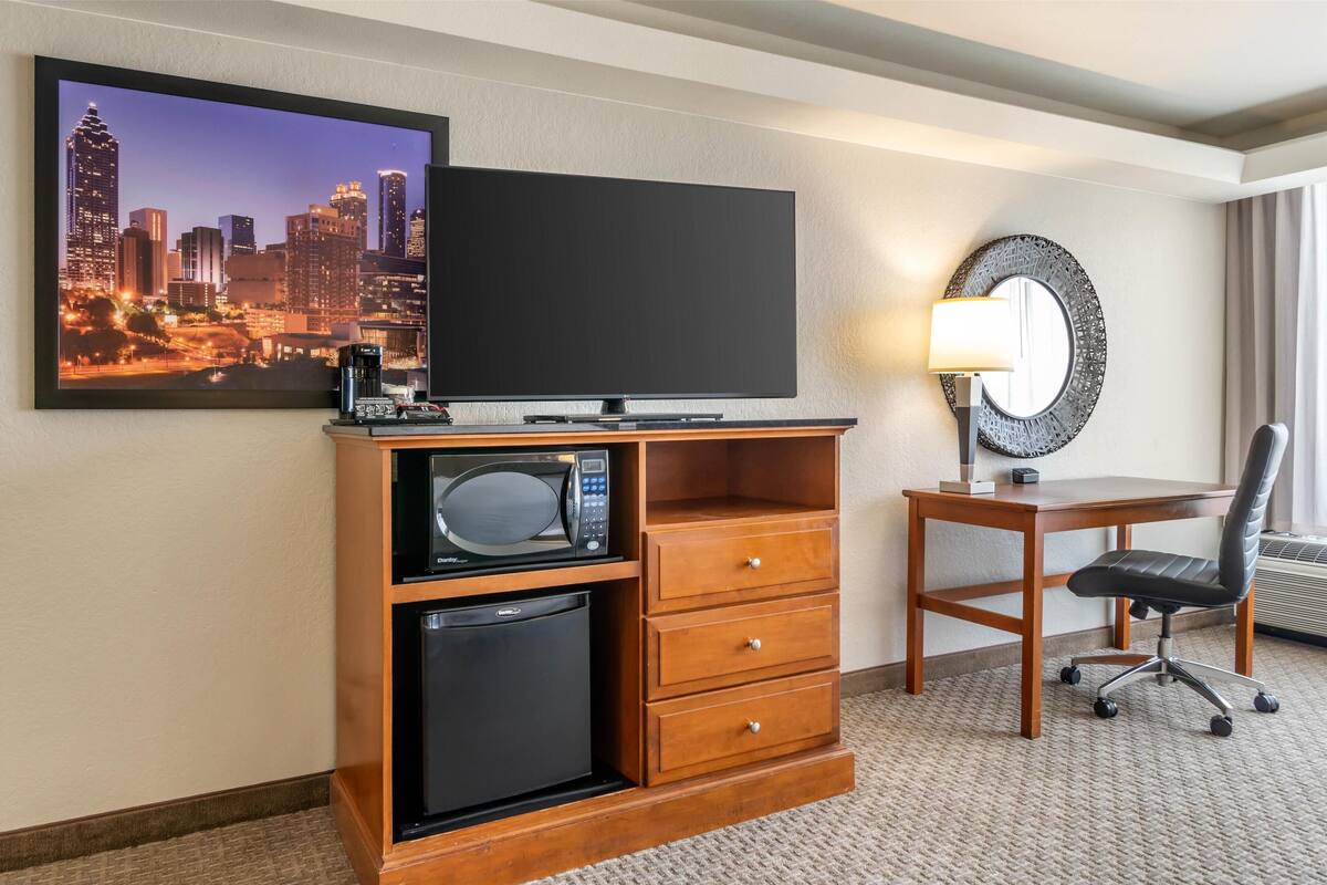 Quarto de hotel com televisão de tela plana, micro-ondas, geladeira, escrivaninha de madeira, cadeira, luminária e um espelho grande.