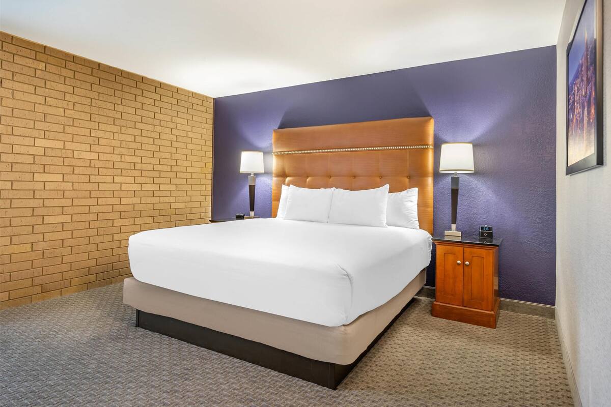 Quarto de hotel com uma cama grande, duas mesas de cabeceira com luminárias, uma mesinha de madeira e uma parede de tijolos.