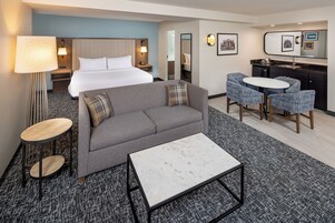 Quarto de hotel com cama, sofá, mesa de centro, escrivaninha e uma pequena cozinha.