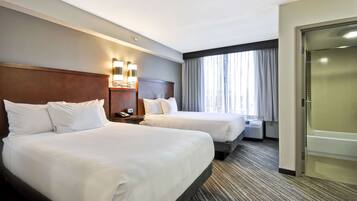 Quarto de hotel com duas camas, uma mesa de cabeceira com telefone, luminária na parede e uma janela grande com cortinas.