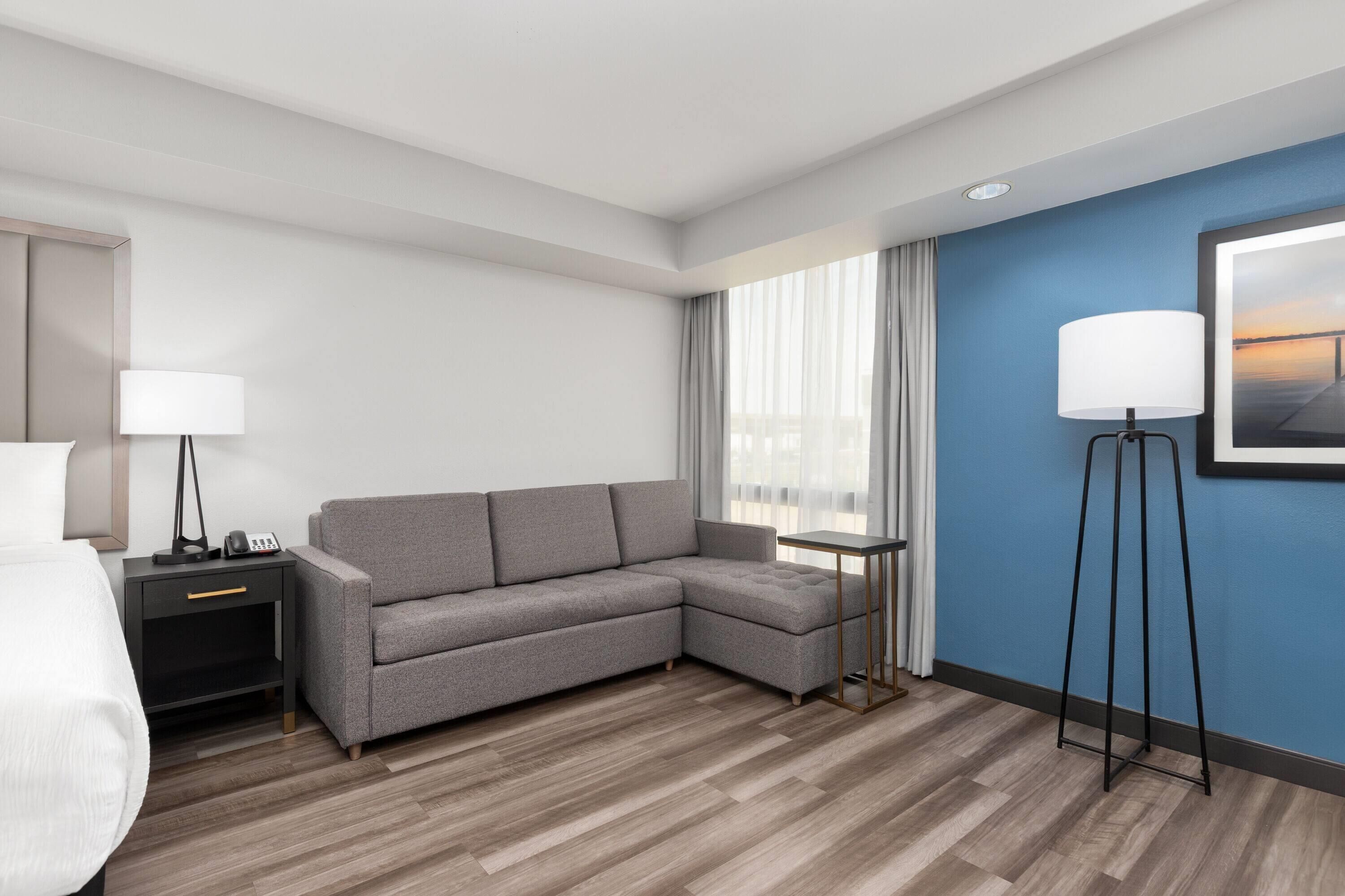 Quarto de hotel moderno com sofá seccional cinza, mesa de cabeceira com luminária, pequena mesa com luminária e um quadro na parede.