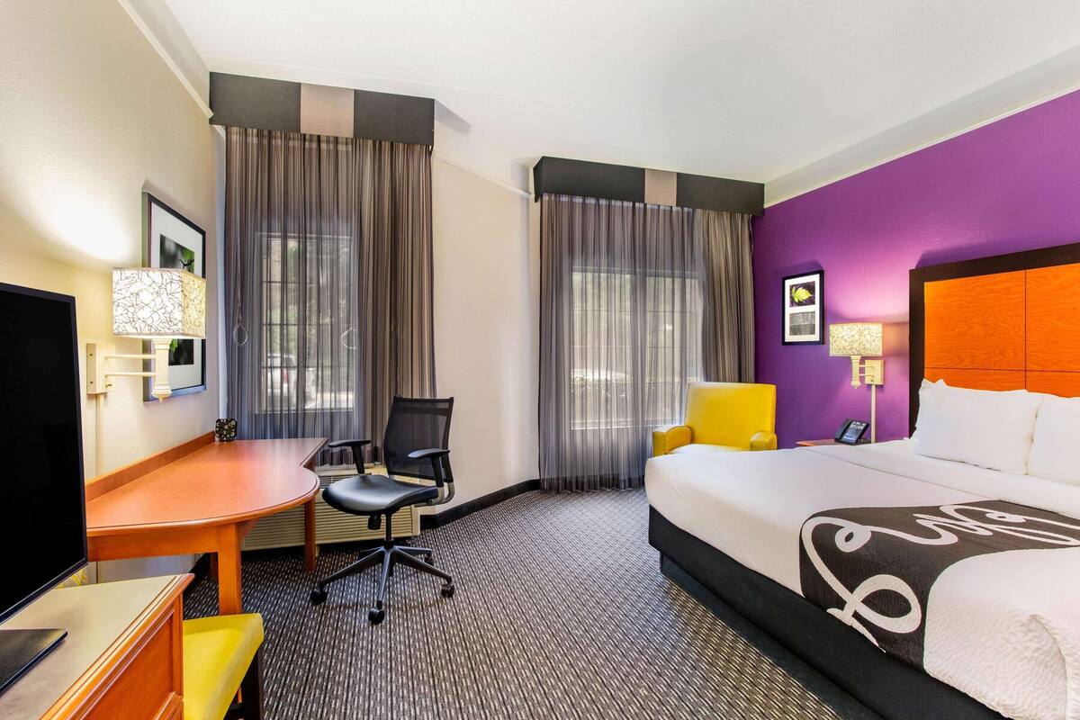 Quarto de hotel com uma cama, uma mesa, uma cadeira, uma televisão e cortinas.