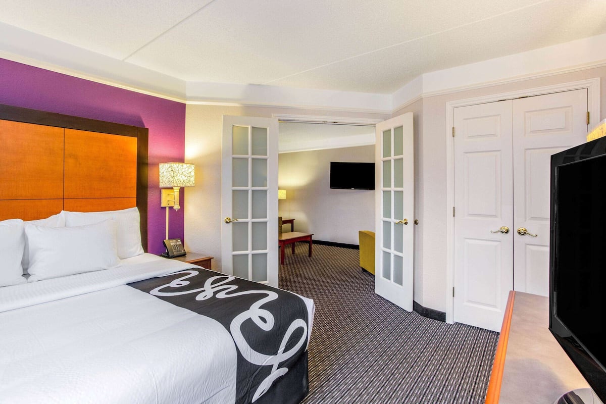 Quarto de hotel com uma cama grande, televisão, secretária com candeeiro e porta para outro espaço.