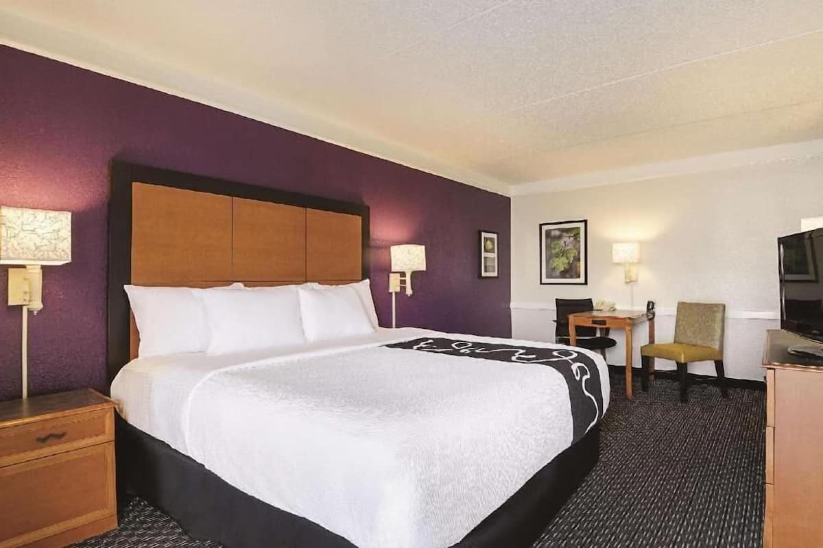 Quarto de hotel com cama, mesas de cabeceira, escrivaninha, cadeira, televisão e luminárias fixadas na parede.