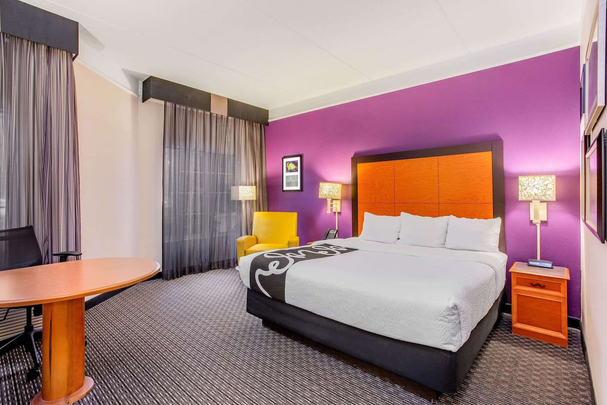 Quarto de hotel com uma cama grande, uma mesa redonda, uma poltrona amarela e uma mesa de cabeceira com abajur.