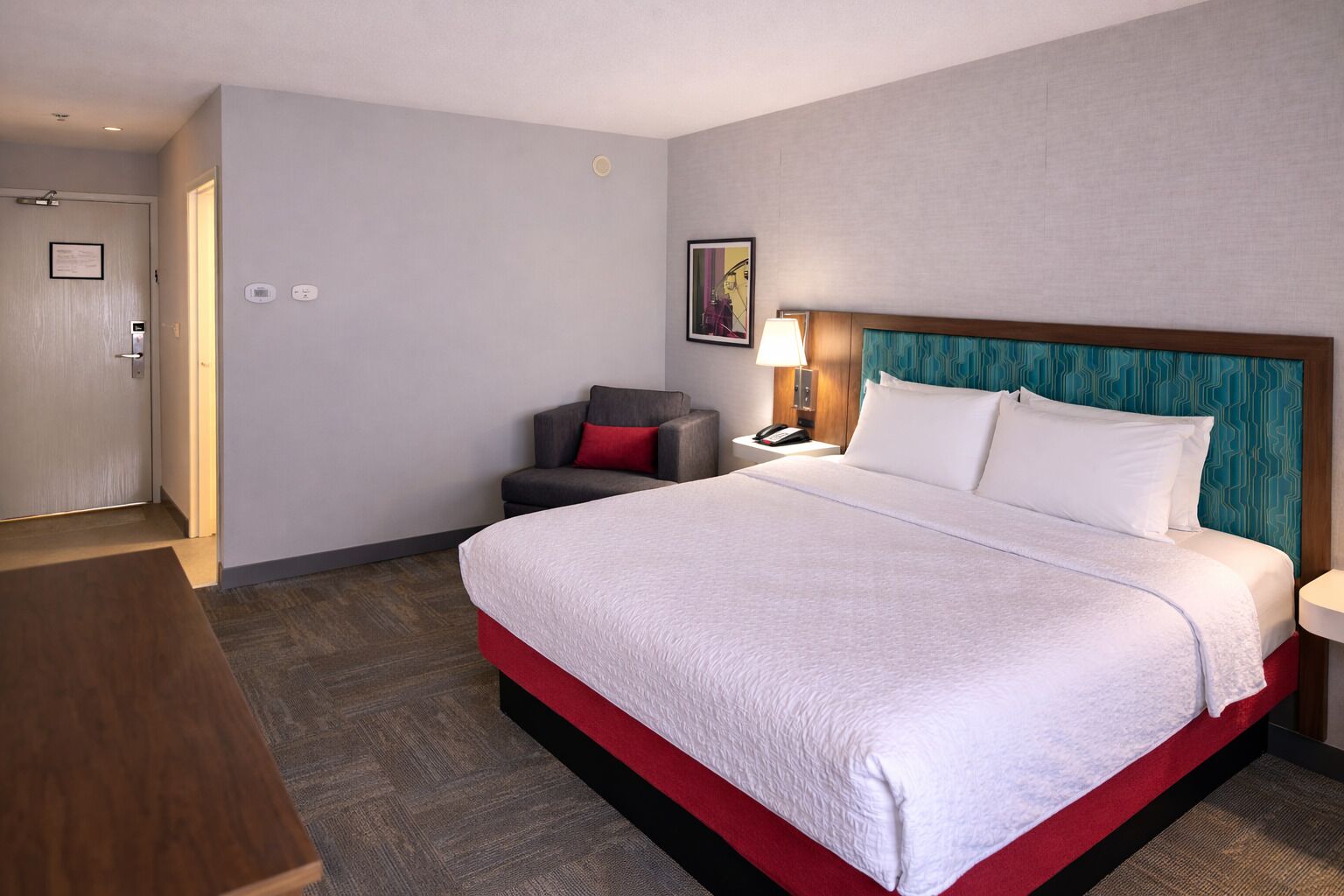 Quarto de hotel com uma cama grande, uma escrivaninha, uma cadeira, uma mesa de cabeceira com abajur e uma porta.