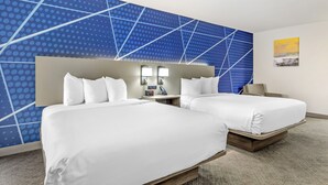 Quarto de hotel com duas camas, um design moderno na parede azul, uma mesinha de cabeceira com abajur e uma pequena pintura na parede.