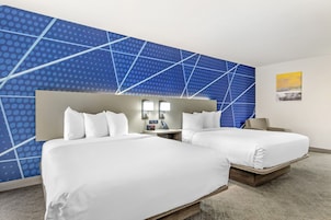 Quarto de hotel com duas camas, um design moderno na parede azul, uma mesinha de cabeceira com abajur e uma pequena pintura na parede.