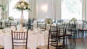 Uma sala de jantar formal com mesas redondas, cadeiras de madeira escura e toalhas de mesa brancas.
