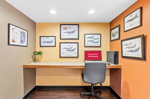 Quarto com escrivaninha, quadros de aviões, uma planta e placa indicando Wi-Fi gratuito.