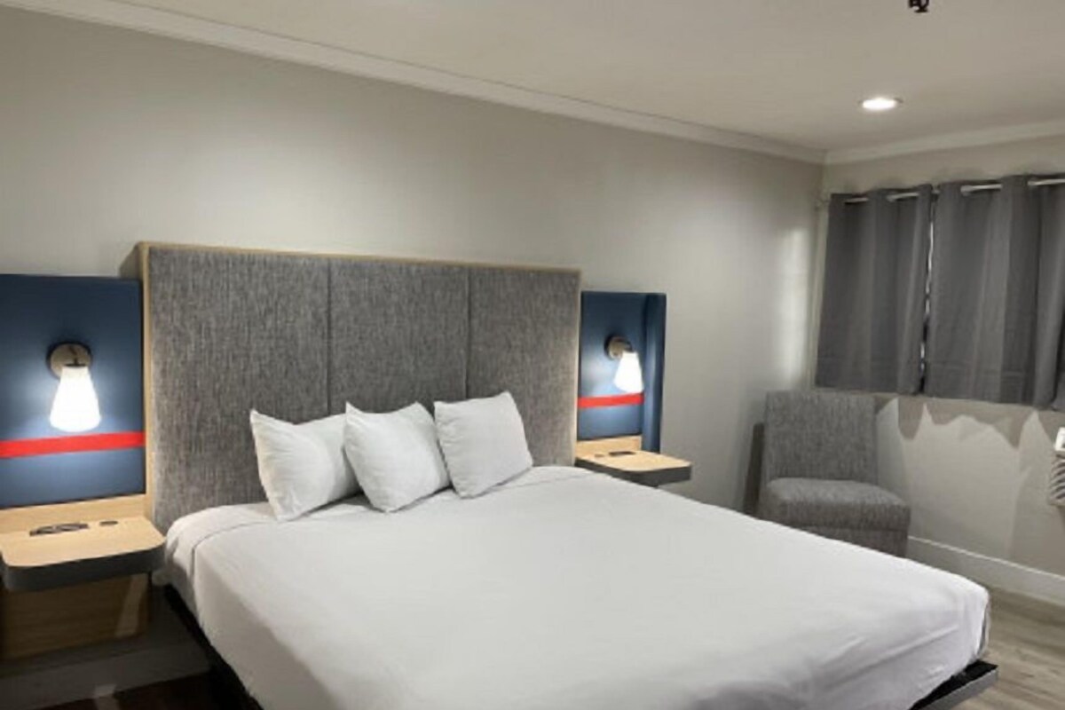 Quarto de hotel com cama grande, duas mesinhas de cabeceira, uma cadeira e janela com cortinas.