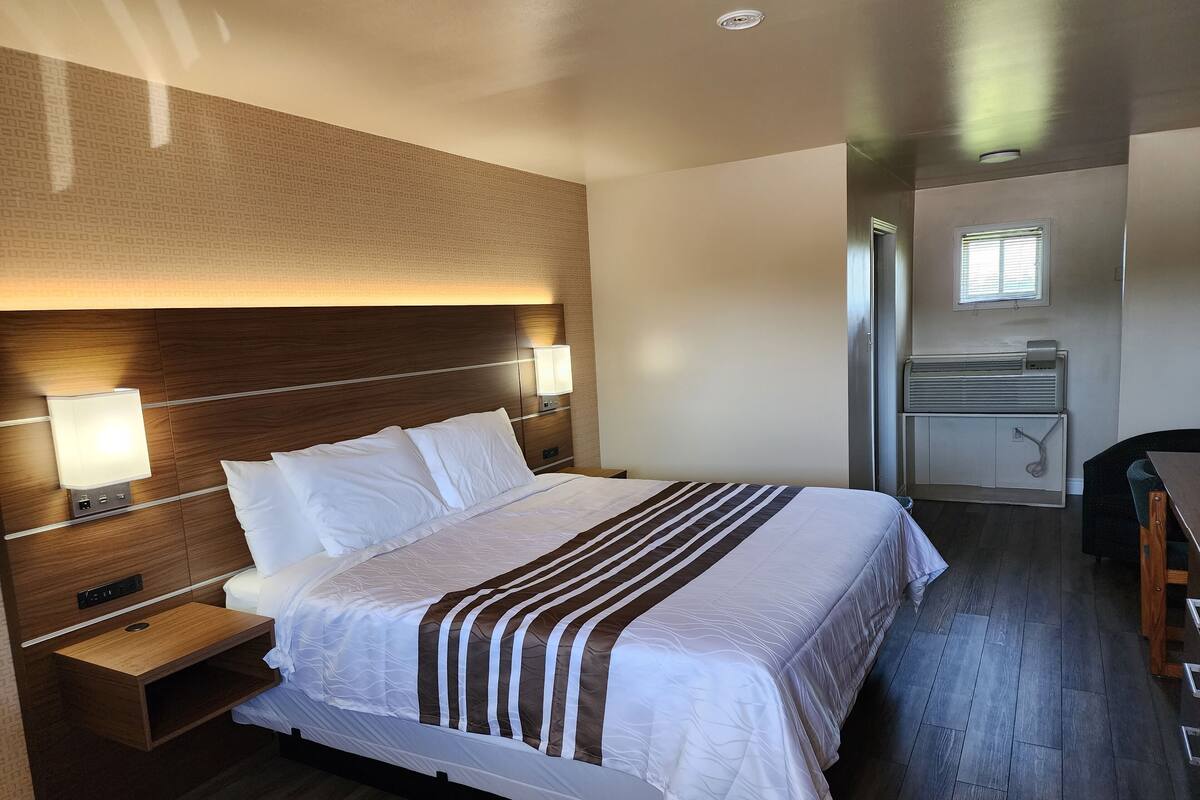 Quarto de hotel com uma cama grande, mesas de cabeceira, uma escrivaninha e uma cozinha compacta.
