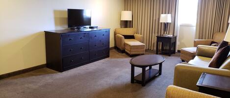 Quarto de hotel com televisão de tela plana em uma cômoda, duas poltronas, um sofá, uma mesa pequena e um abajur.