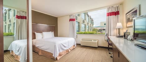Quarto de hotel com uma cama grande, uma escrivaninha, uma cadeira e uma televisão. Há uma janela com cortinas e vista para edifícios.