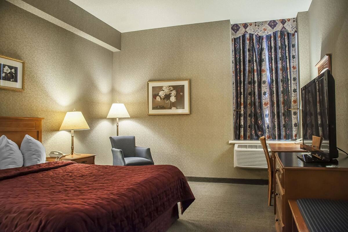 Quarto de hotel com cama, duas luminárias de cabeceira, uma cadeira, escrivaninha com computador, quadro emoldurado e janela com cortinas.