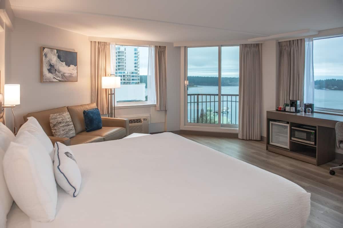 Quarto de hotel com uma cama grande, uma escrivaninha com computador, uma cadeira, um abajur e vista para um corpo de água por grandes janelas.