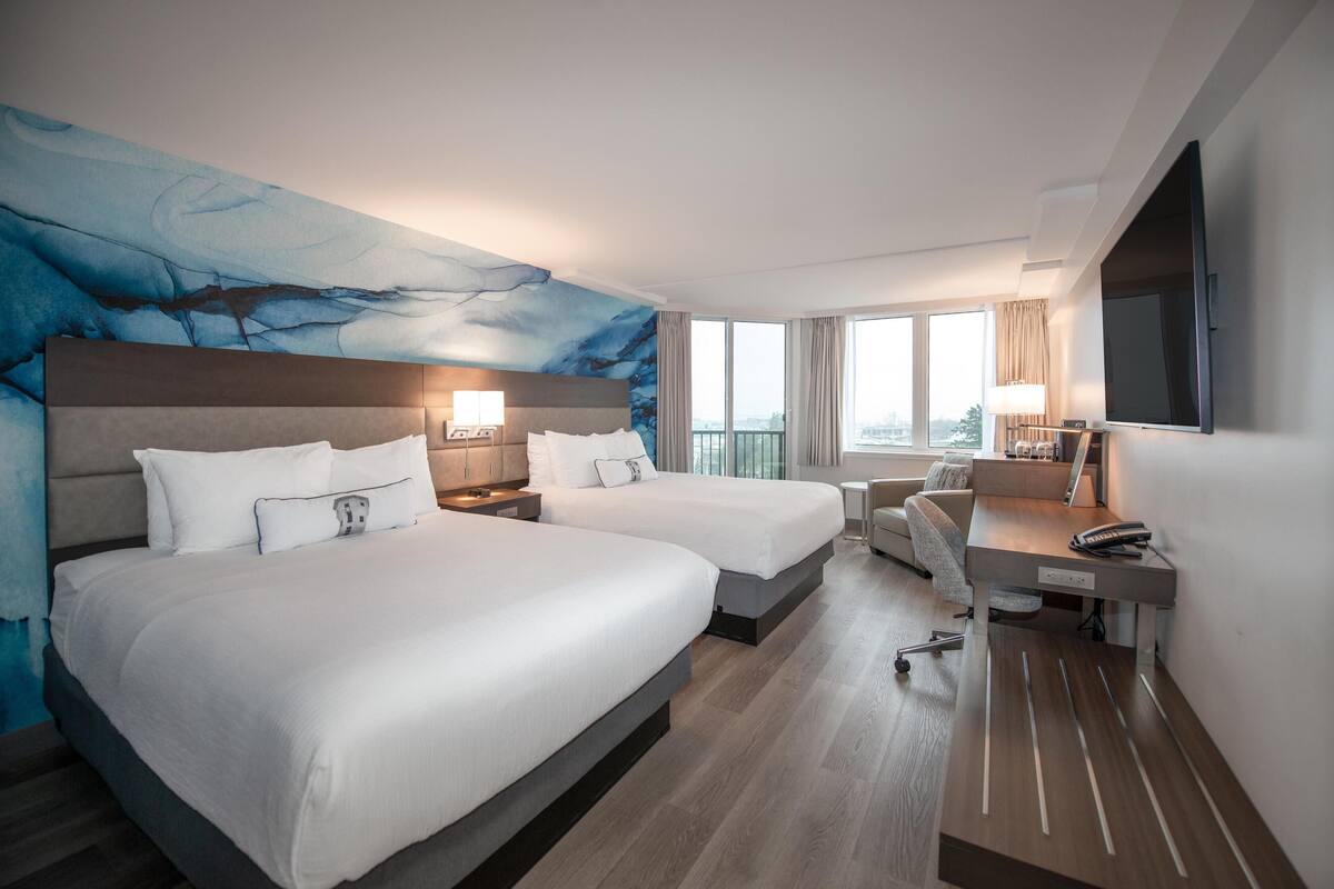 Quarto de hotel com duas camas, uma escrivaninha, televisão e uma janela grande.