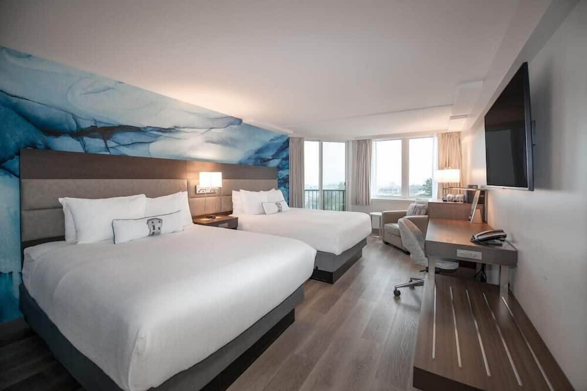 Quarto de hotel com duas camas, uma escrivaninha, televisão e uma janela grande.