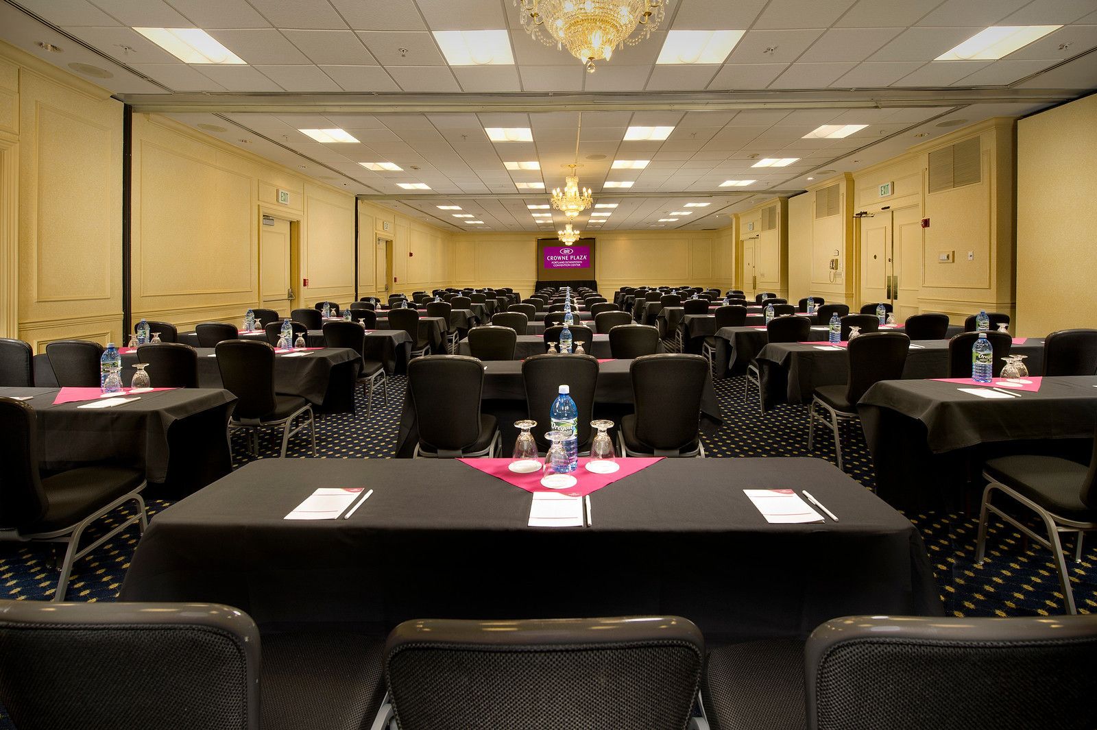 Uma sala de conferências com fileiras de mesas e cadeiras, equipada com garrafas de água e placas de identificação.